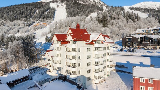 Um hotel com vários andares e teto vermelho, rodeado por árvores e montanhas cobertas de neve.