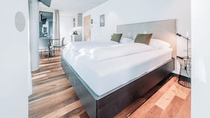 Quarto de hotel moderno com uma cama grande, piso de madeira, escrivaninha e uma cozinha compacta.