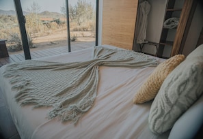 Uma cama com cobertor, travesseiros e vista para uma paisagem desértica através de grandes janelas.