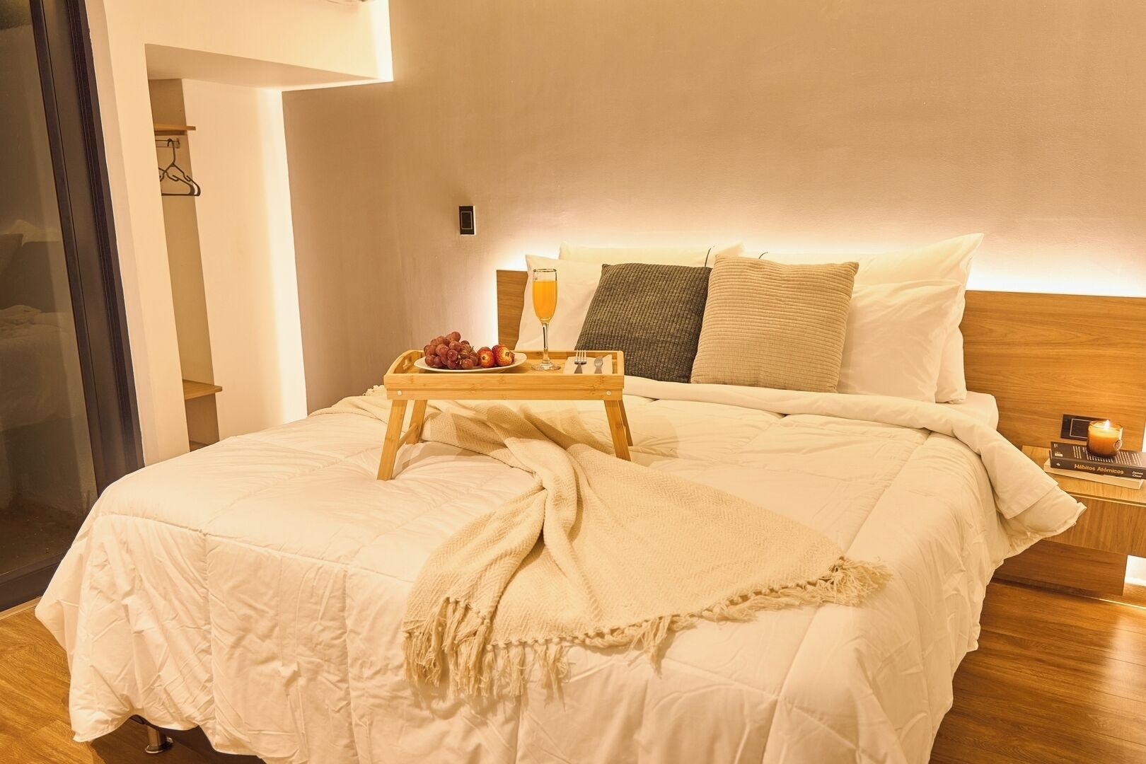 Cama bem arrumada com bandeja de frutas e um copo de suco de laranja, mesa de cabeceira com uma vela e cabeceira de madeira.