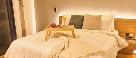 Cama bem arrumada com bandeja de frutas e um copo de suco de laranja, mesa de cabeceira com uma vela e cabeceira de madeira.