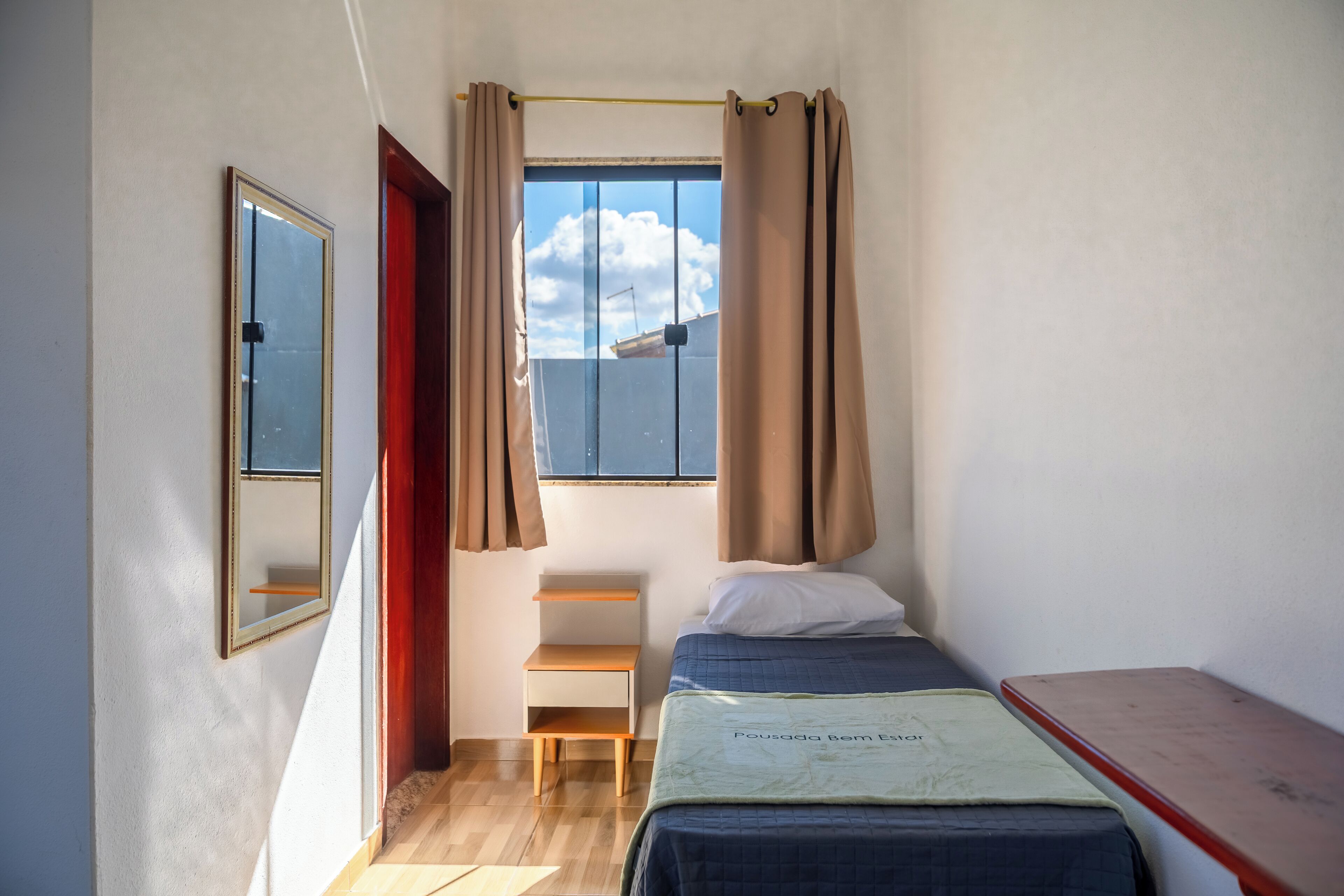 Quarto pequeno e modesto, com cama, janela com cortinas, espelho e pequena prateleira de madeira.