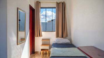Quarto pequeno e modesto, com cama, janela com cortinas, espelho e pequena prateleira de madeira.