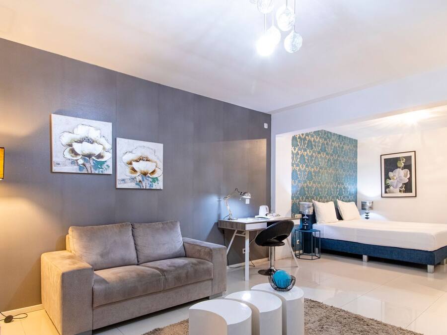 Quarto de hotel moderno com sofá cinzento, secretária, cama com cabeceira azul e obras de arte nas paredes.
