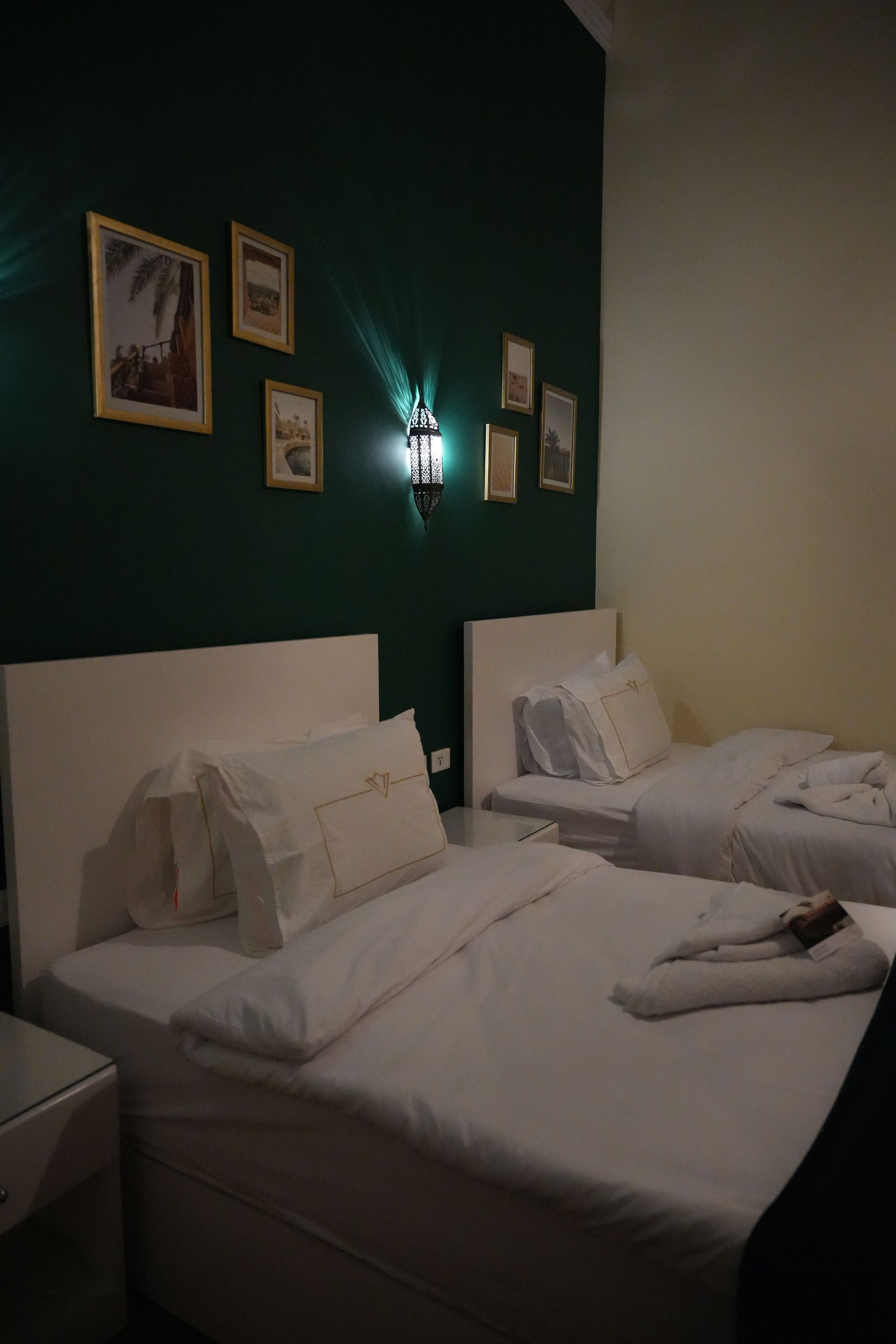 Quarto de hotel com duas camas, uma mesinha de cabeceira e um ponto de luz fixado na parede.