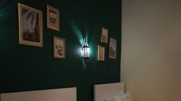 Quarto de hotel com duas camas, uma mesinha de cabeceira e um ponto de luz fixado na parede.
