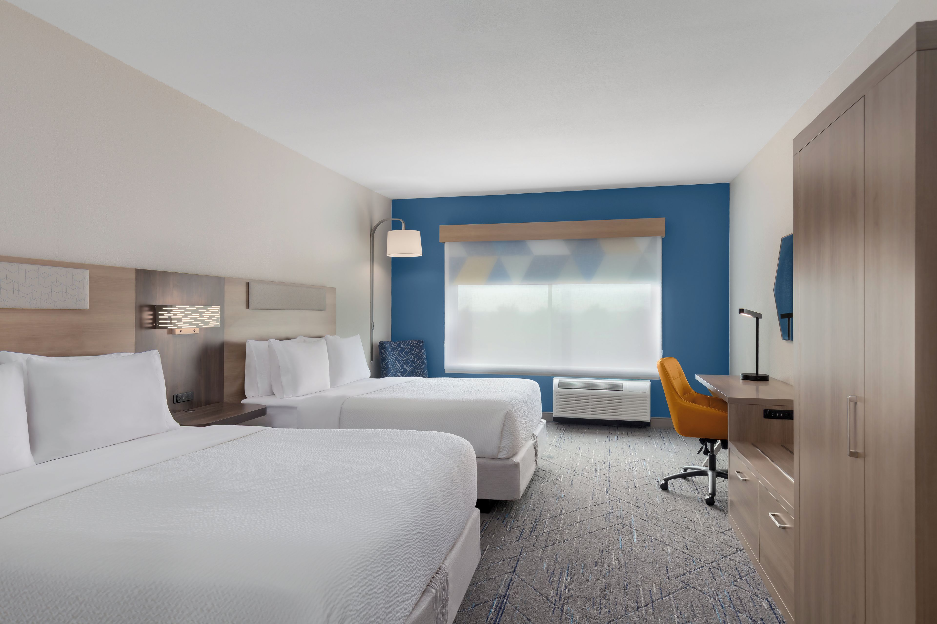 Quarto de hotel com duas camas, uma escrivaninha, uma cadeira, janela com persianas e ar condicionado fixado na parede.