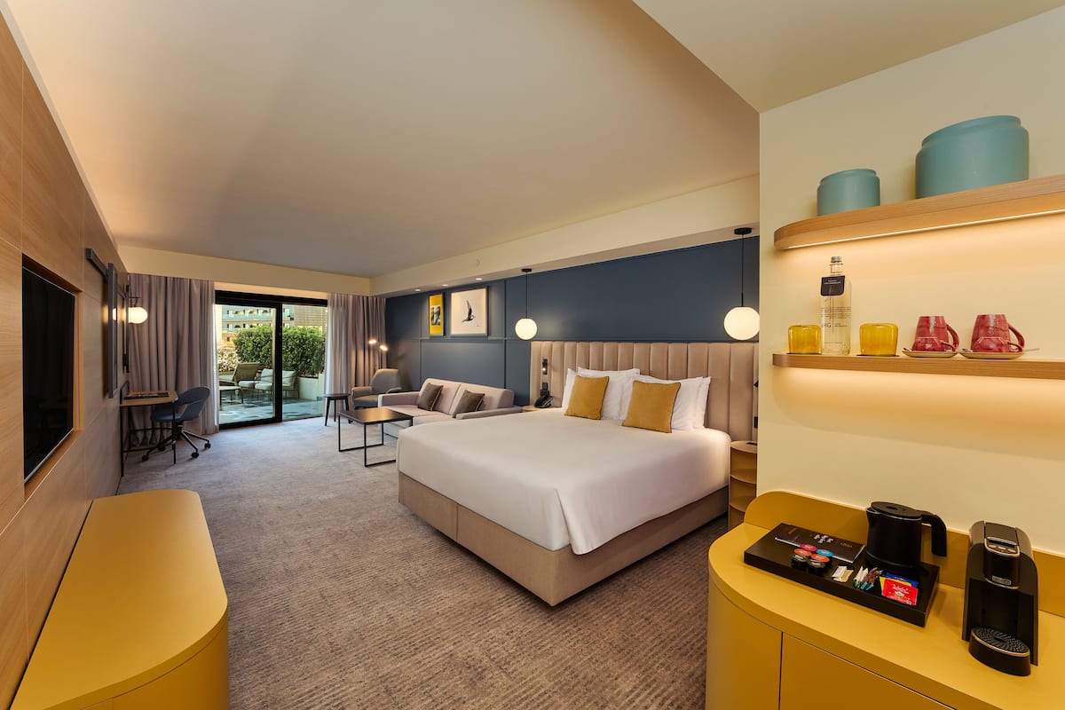 Quarto de hotel moderno com uma cama grande, escrivaninha, sofá, cantinho do café e vista para o exterior.