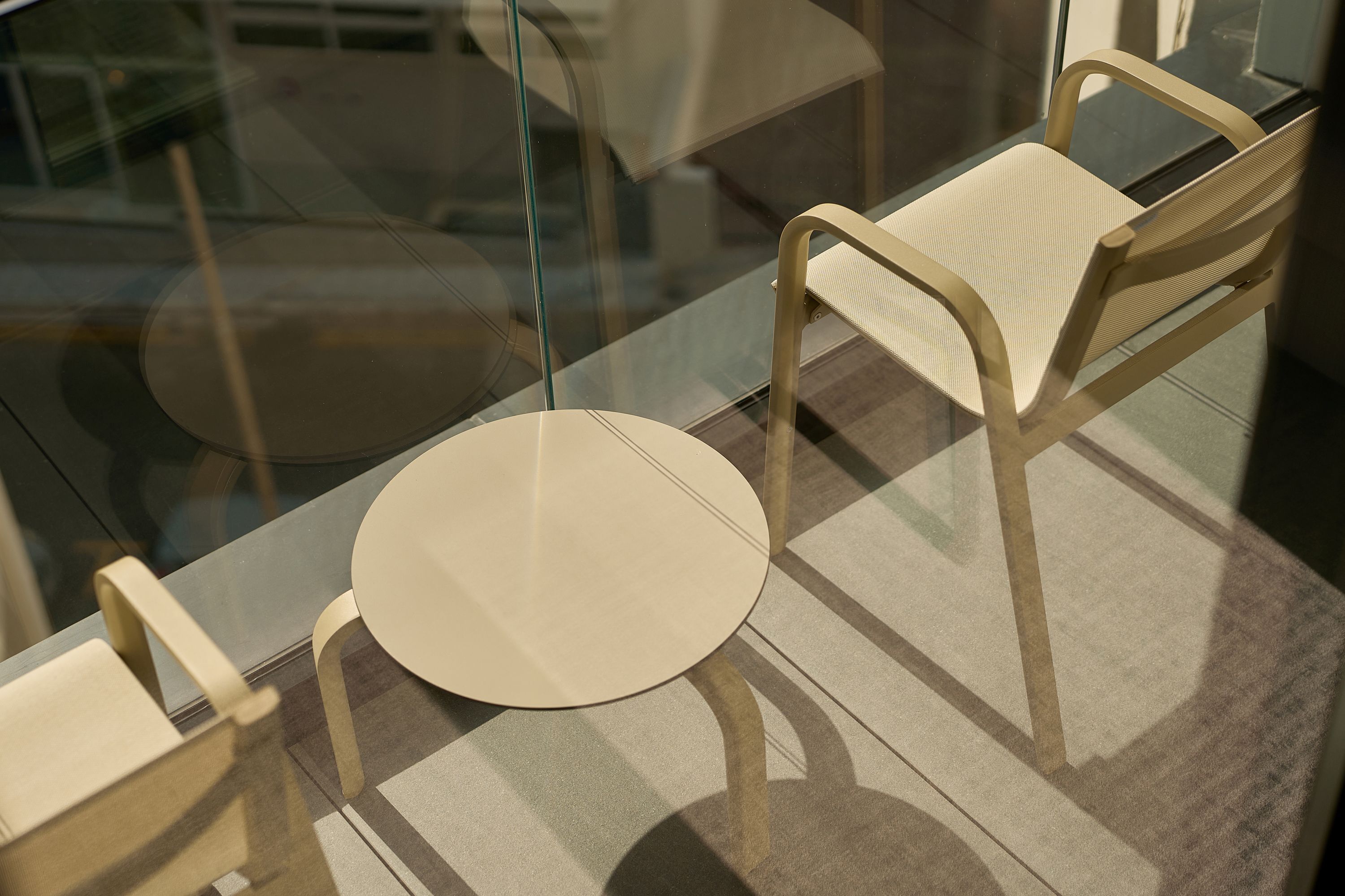 Uma mesa de vidro com tampo redondo branco, duas cadeiras bege com assentos trançados e um painel de vidro.