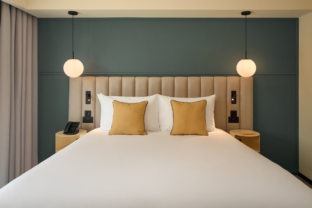 Quarto de hotel com uma cama grande, duas mesas de cabeceira, um telefone na mesa da esquerda e um candeeiro fixo na parede.