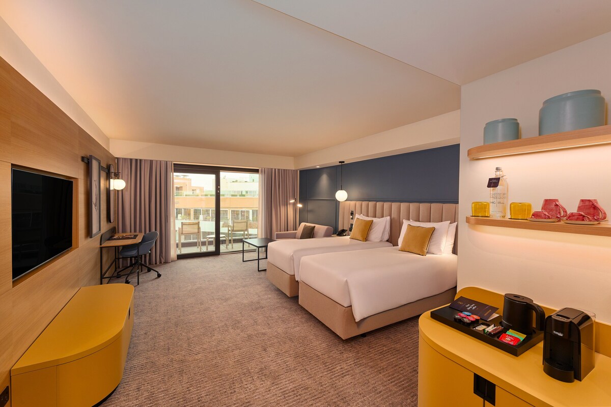 Quarto de hotel com duas camas, uma escrivaninha, televisão e varanda com vista.