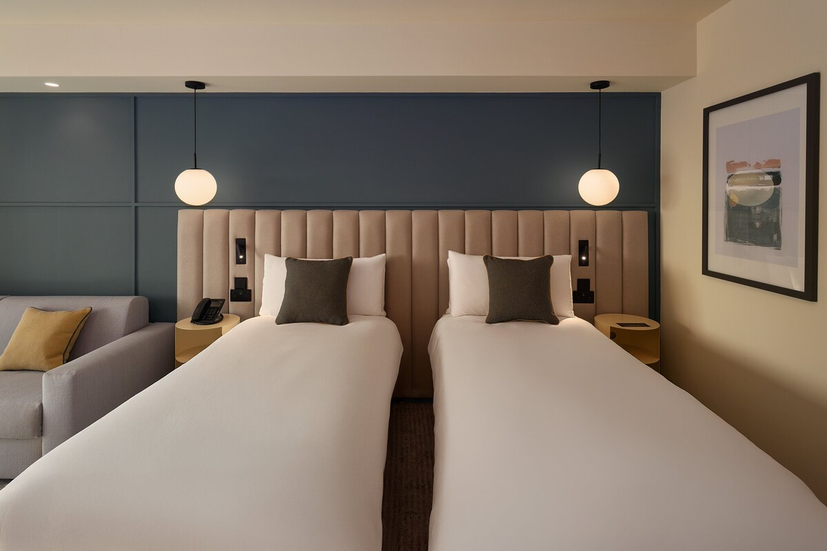 Quarto de hotel com duas camas, um sofá, uma mesa de cabeceira com telefone e uma obra de arte na parede.