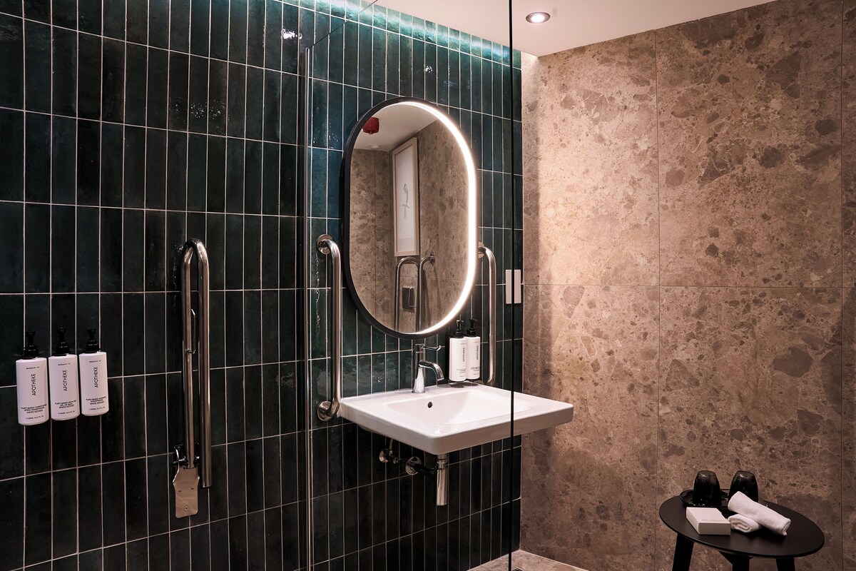 Uma casa de banho moderna com um espelho grande, uma pia e barras de apoio. Há artigos de higiene pessoal na parede e uma toalha sobre uma mesinha.