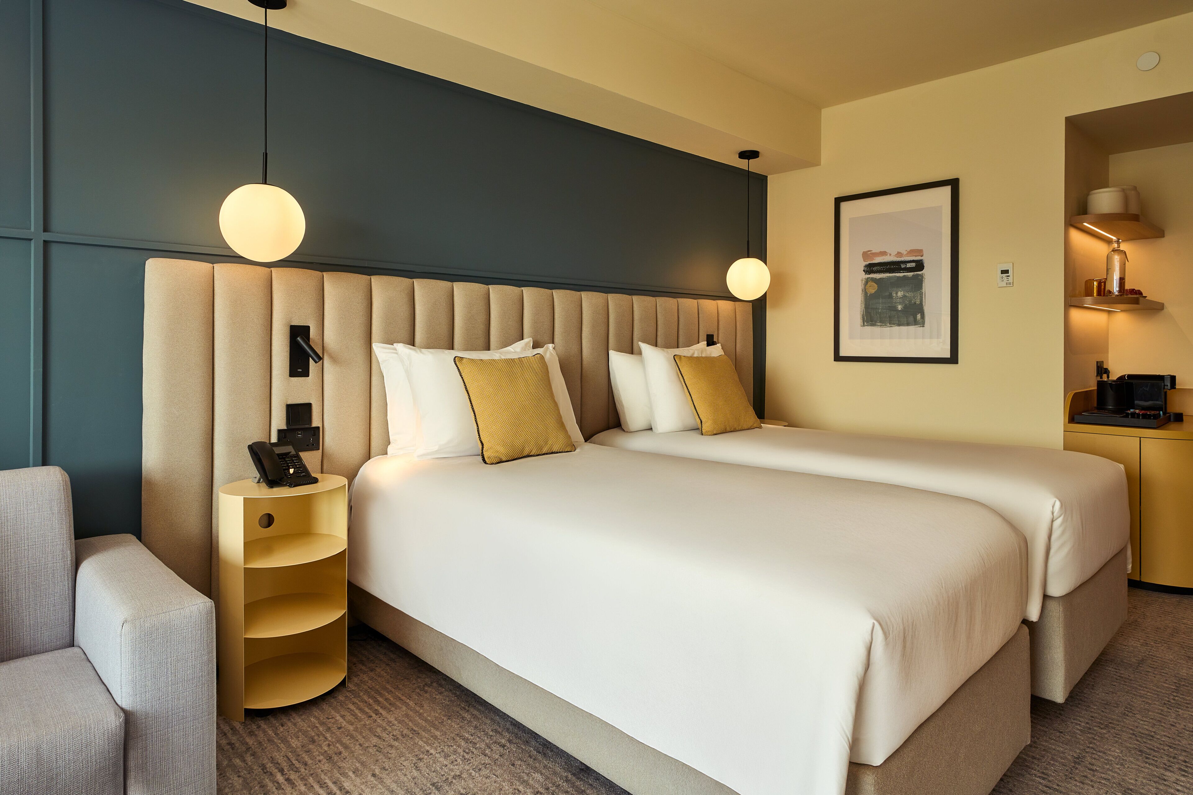 Quarto de hotel com duas camas, uma escrivaninha, uma cadeira e um abajur.