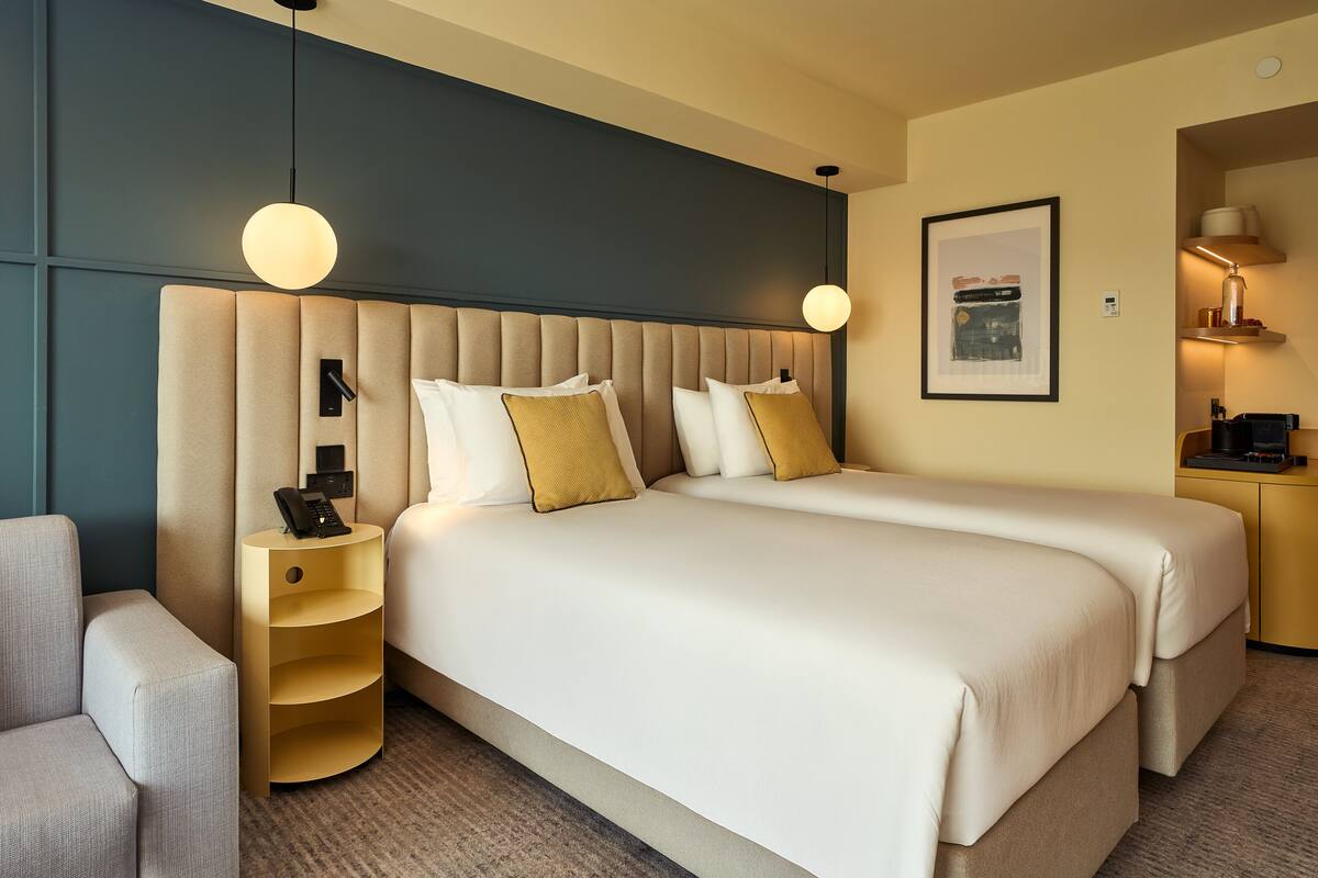 Quarto de hotel com duas camas, uma escrivaninha, uma cadeira e um abajur.
