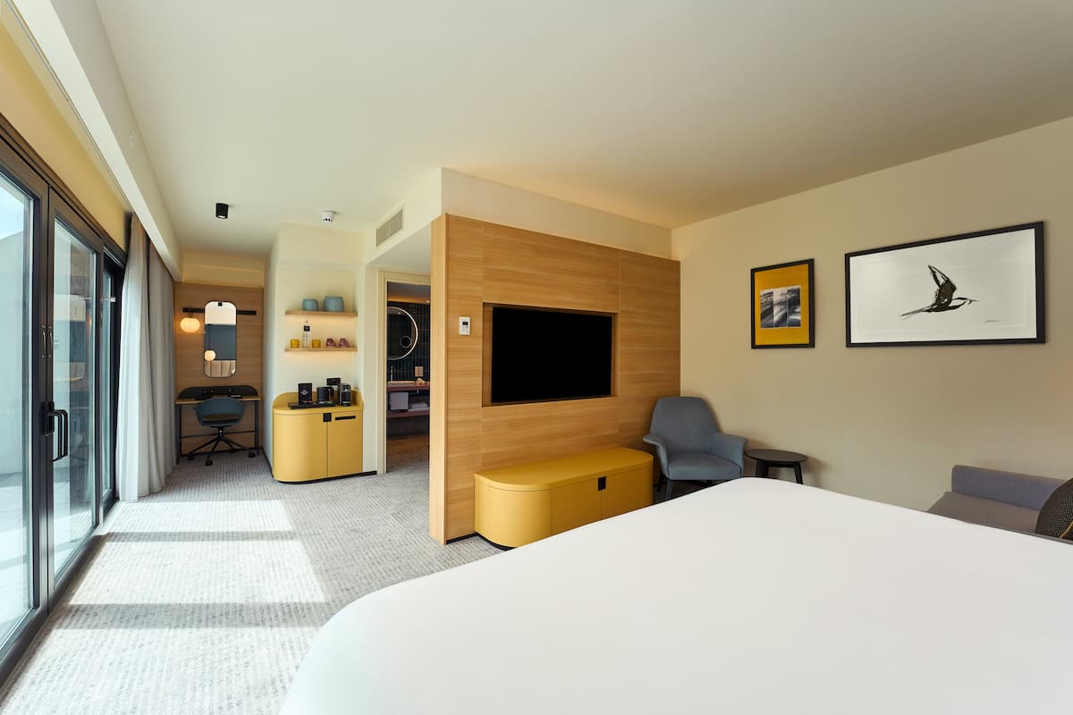 Quarto de hotel com uma cama grande, uma escrivaninha, uma cadeira, televisão e obras de arte nas paredes.