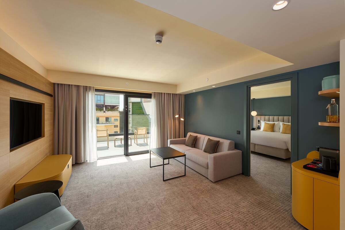 Quarto de hotel moderno com sofá, mesa pequena, cama, escrivaninha e uma janela grande com cortinas.