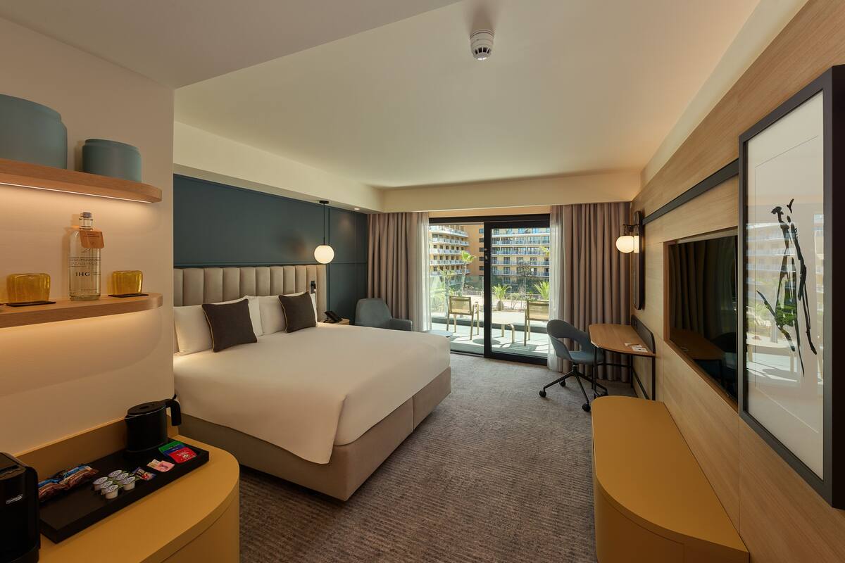 Quarto de hotel com cama, mesa de cabeceira, abajur e vista para um edifício pela janela.