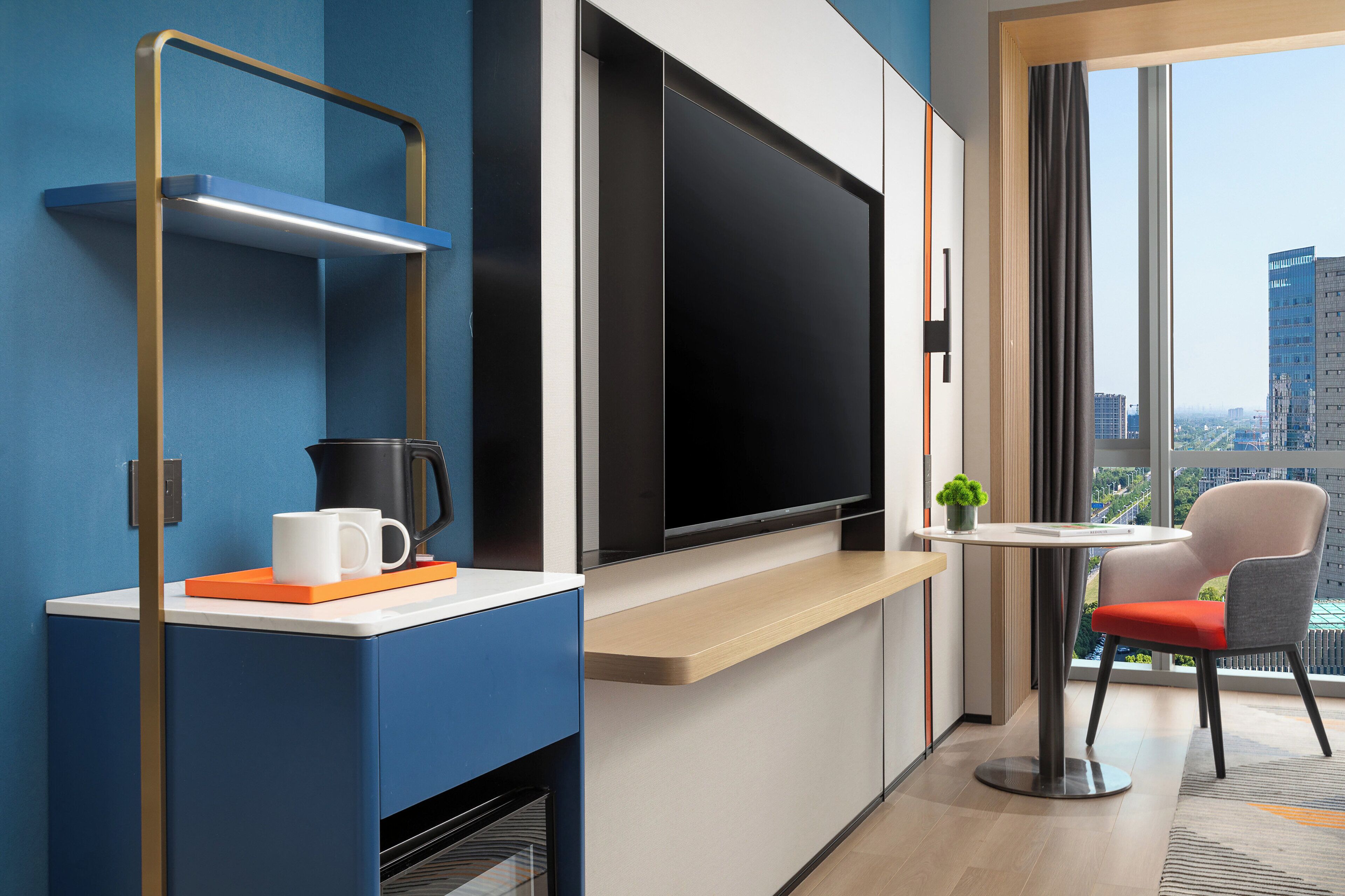 Quarto de hotel moderno com uma janela grande, televisão de tela plana, cafeteira, caneca branca e uma pequena mesa com uma cadeira.
