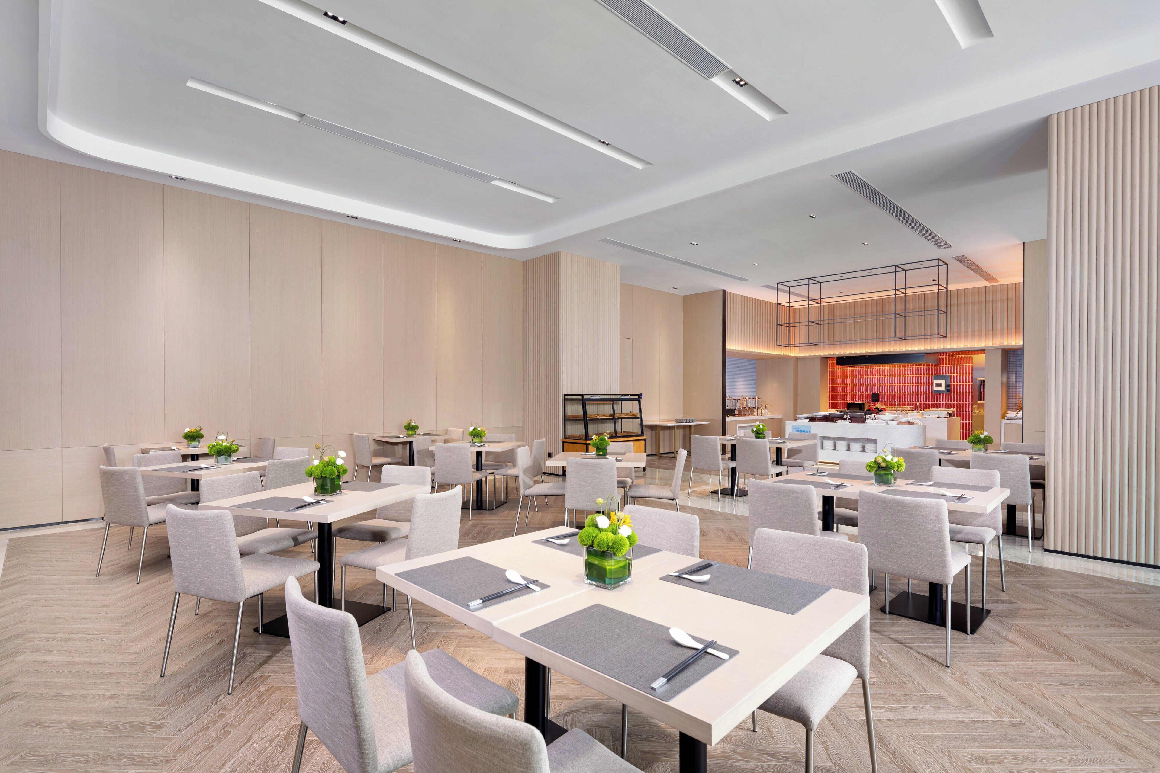 Espaço de refeições moderno em hotel, com várias mesas e cadeiras, cozinha ao fundo e design limpo e minimalista.