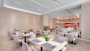 Espaço de refeições moderno em hotel, com várias mesas e cadeiras, cozinha ao fundo e design limpo e minimalista.
