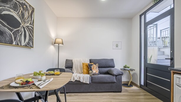 Uma sala de estar moderna com um sofá cinzento, uma mesa de jantar de madeira com café da manhã, um abajur e uma janela grande com vista para o exterior.