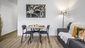 Uma sala de estar moderna com área de jantar, apresentando uma mesa redonda com cadeiras, um sofá com manta e almofadas, um abajur e uma pintura abstrata na parede.