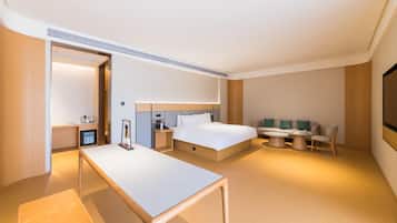 Quarto de hotel moderno com cama, mesa de cabeceira, escrivaninha, cadeira e área de estar com sofá e poltronas.