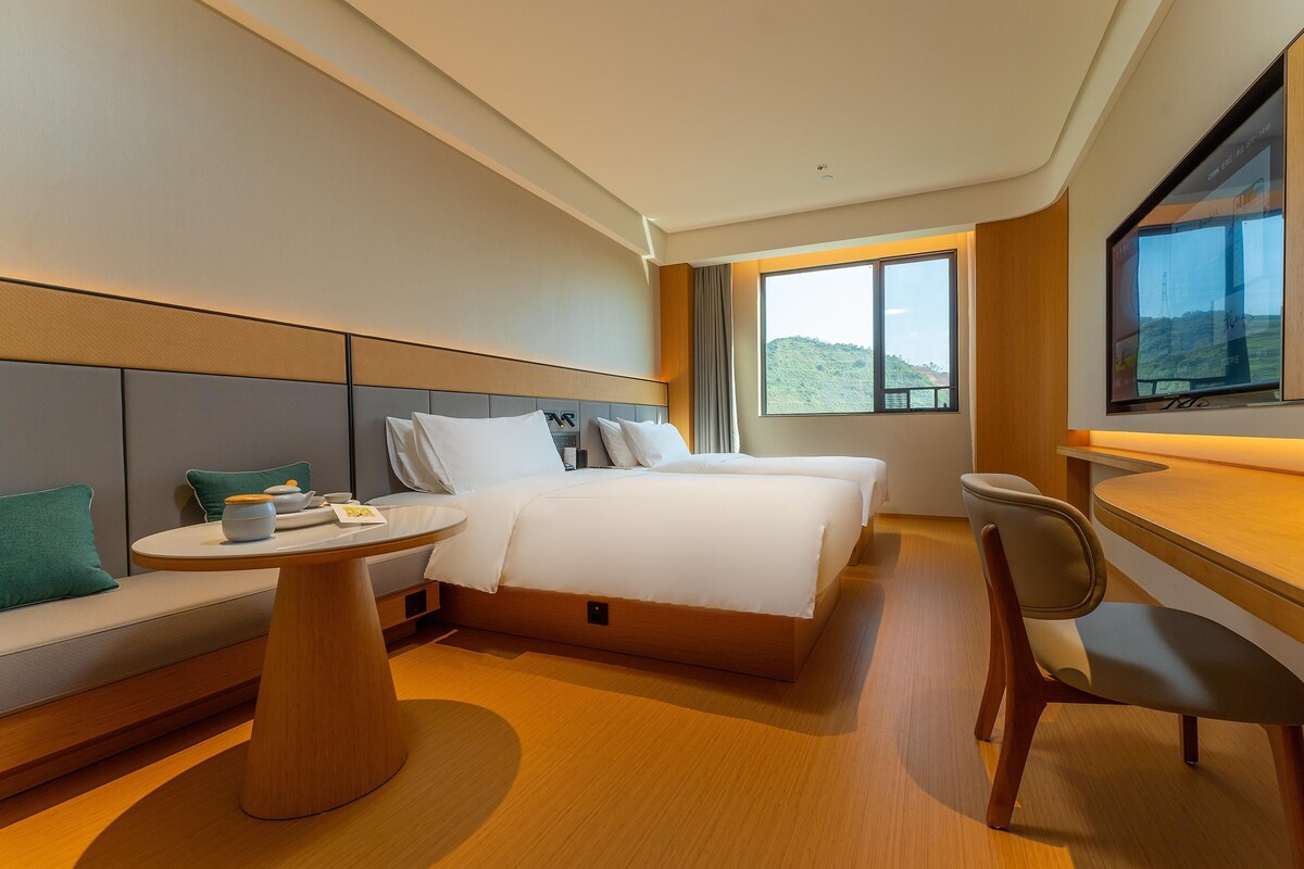 Quarto de hotel moderno com uma cama, uma escrivaninha, uma cadeira, uma mesa redonda e vista para o exterior.