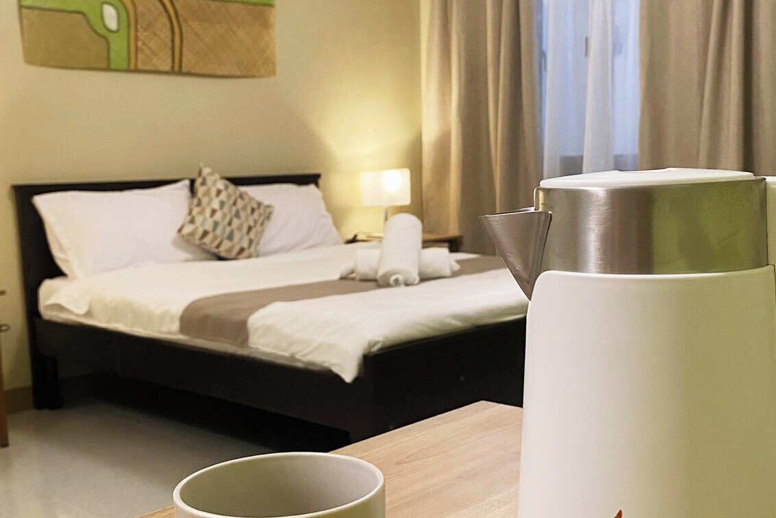 Quarto de hotel com cama, mesa de cabeceira com abajur e cafeteira grande.