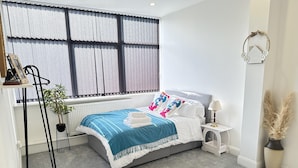 Quarto moderno com carpete cinza, cama com edredom azul e branco, mesa de cabeceira com abajur, radiador e janela com cortinas.