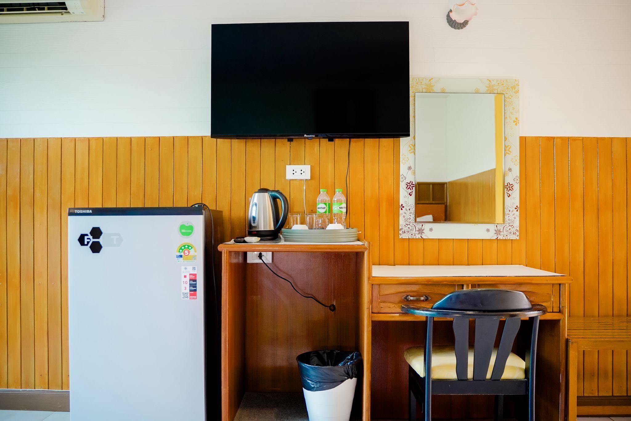 Quarto compacto com geladeira, TV de tela plana na parede, escrivaninha de madeira, cadeira e uma pequena lixeira.