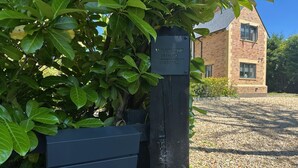 Caixa de eletricidade preta externa com uma placa que diz "WOODSIDE HOUSE OX331A" fixada a um poste de madeira, cercada por folhagem verde.