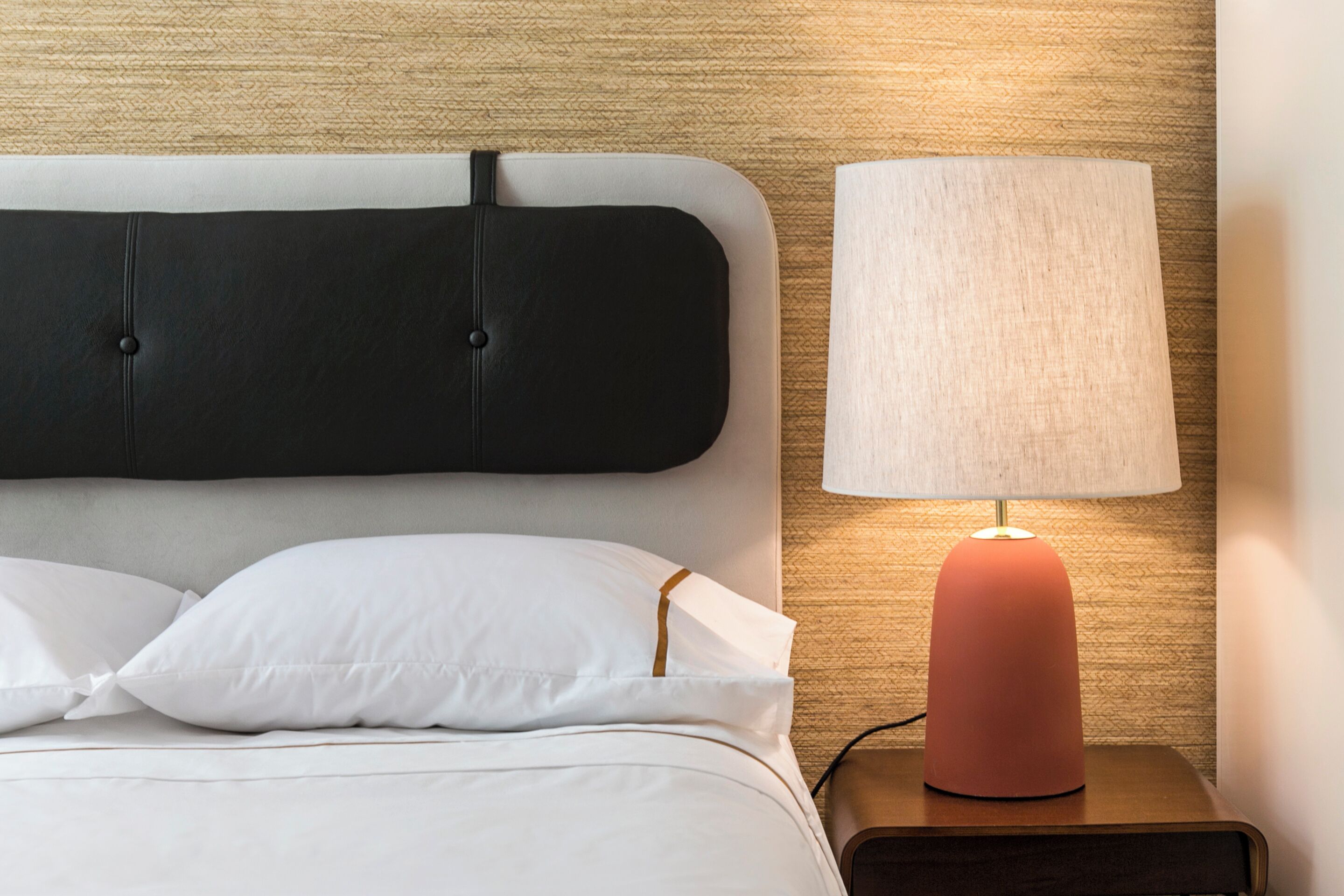 Uma cama bem arrumada com roupa de cama branca e cabeceira preta, acompanhada de uma mesa de cabeceira com abajur rosa.
