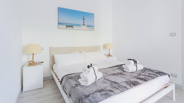 Quarto de hotel com uma cama, duas mesas de cabeceira com luminárias, um quadro com uma cena de praia e um banquinho.