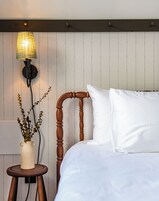 Uma cama bem arrumada com lençóis brancos, cabeceira de madeira, luminária fixada na parede e um pequeno banquinho de madeira com um vaso de flores secas.