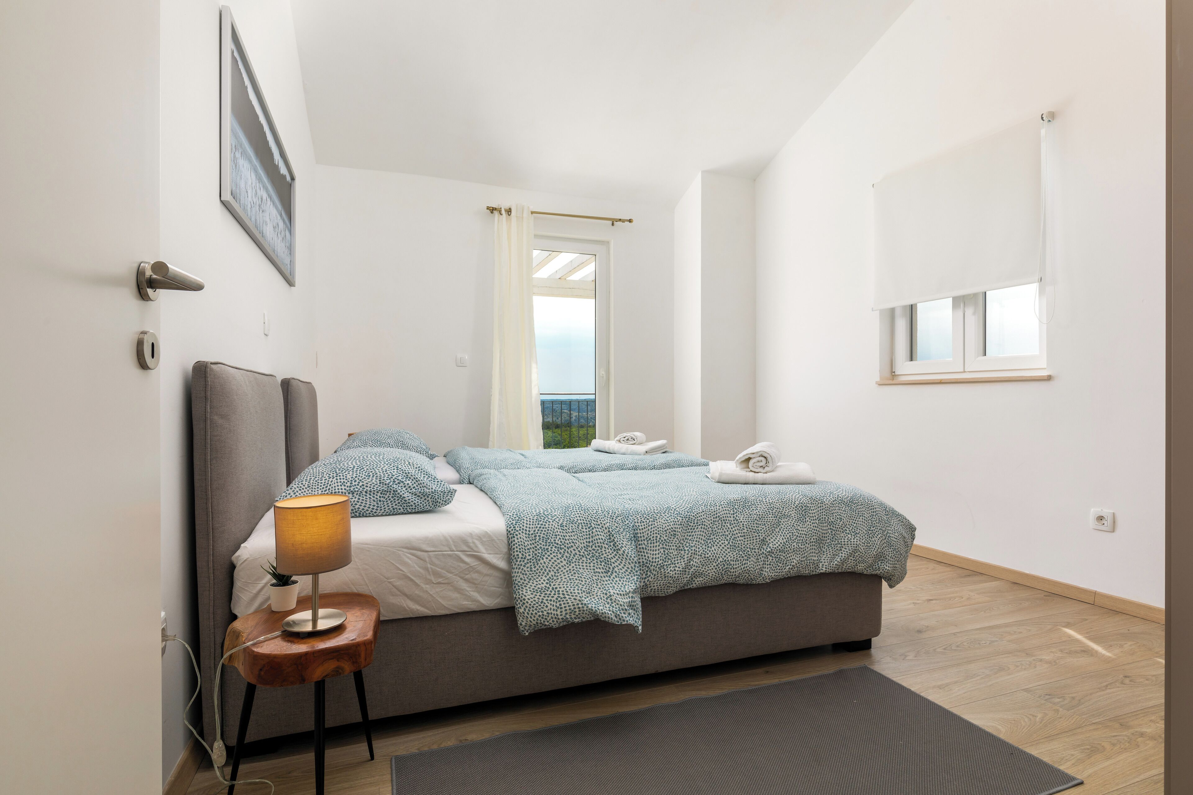 Quarto com cama, mesa de cabeceira, abajur e vista para o exterior.