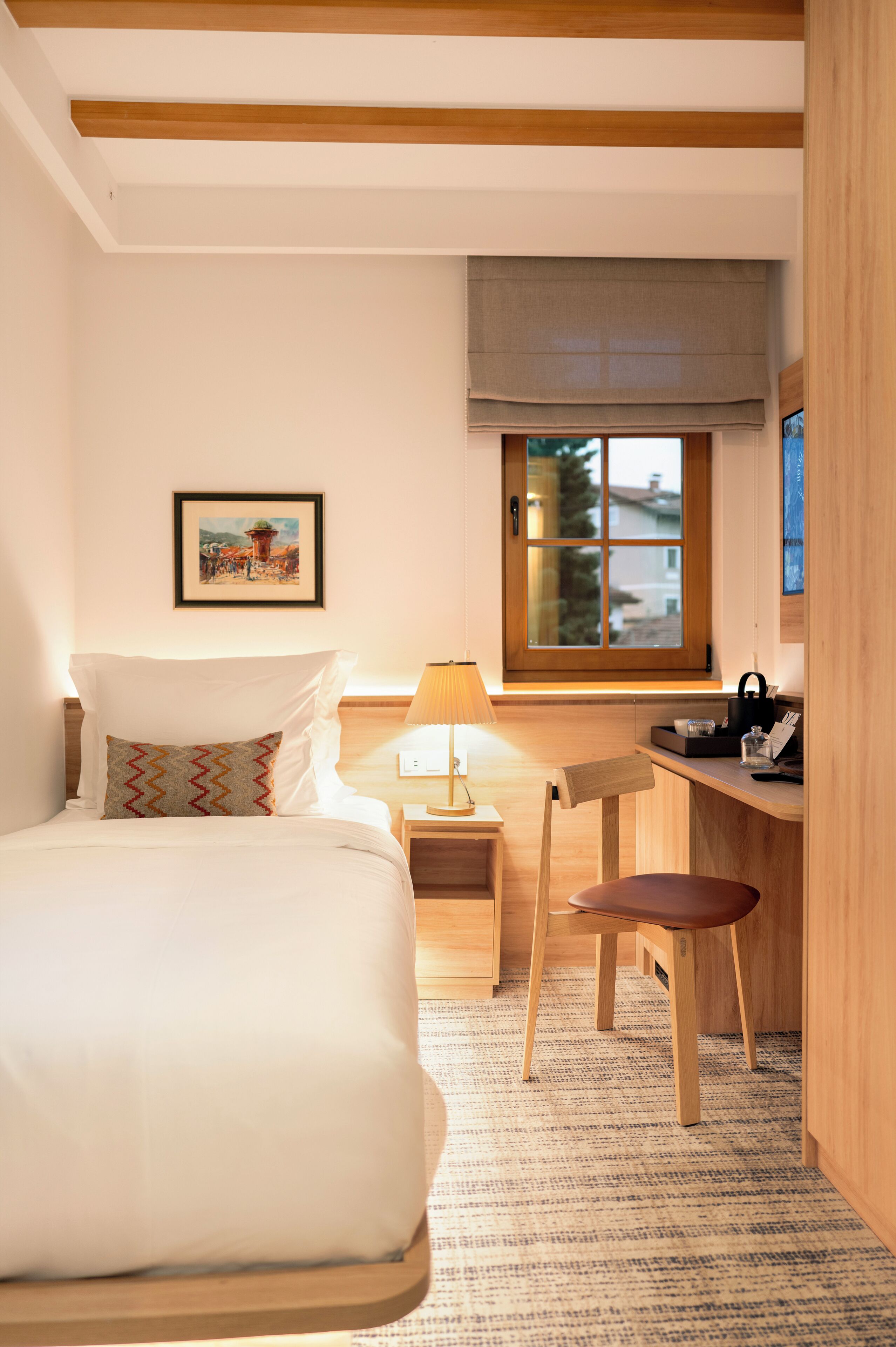Quarto de hotel com cama, escrivaninha, cadeira, abajur, janela e obras de arte.