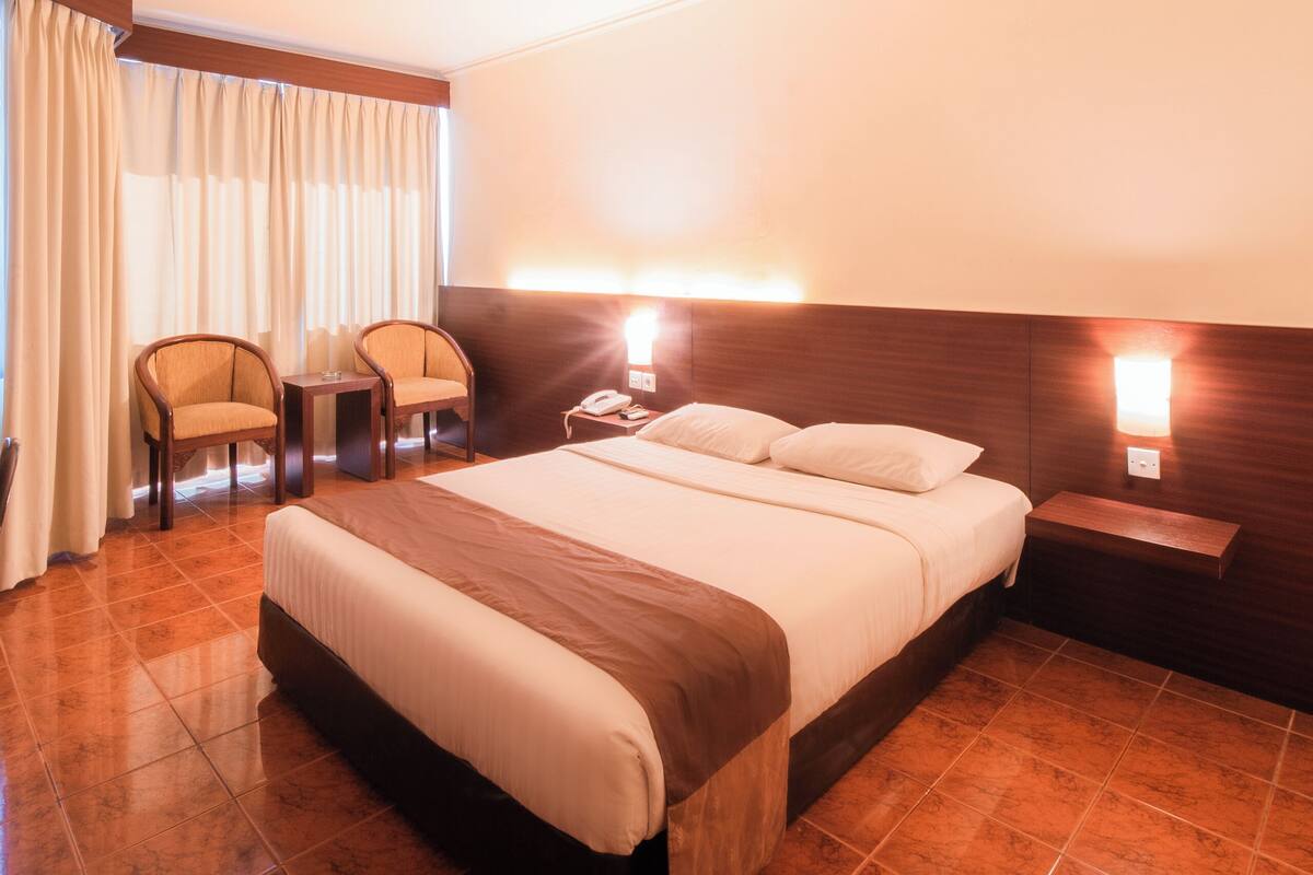 Quarto de hotel com uma cama grande, duas poltronas, uma mesinha, um criado-mudo e uma janela com cortinas.