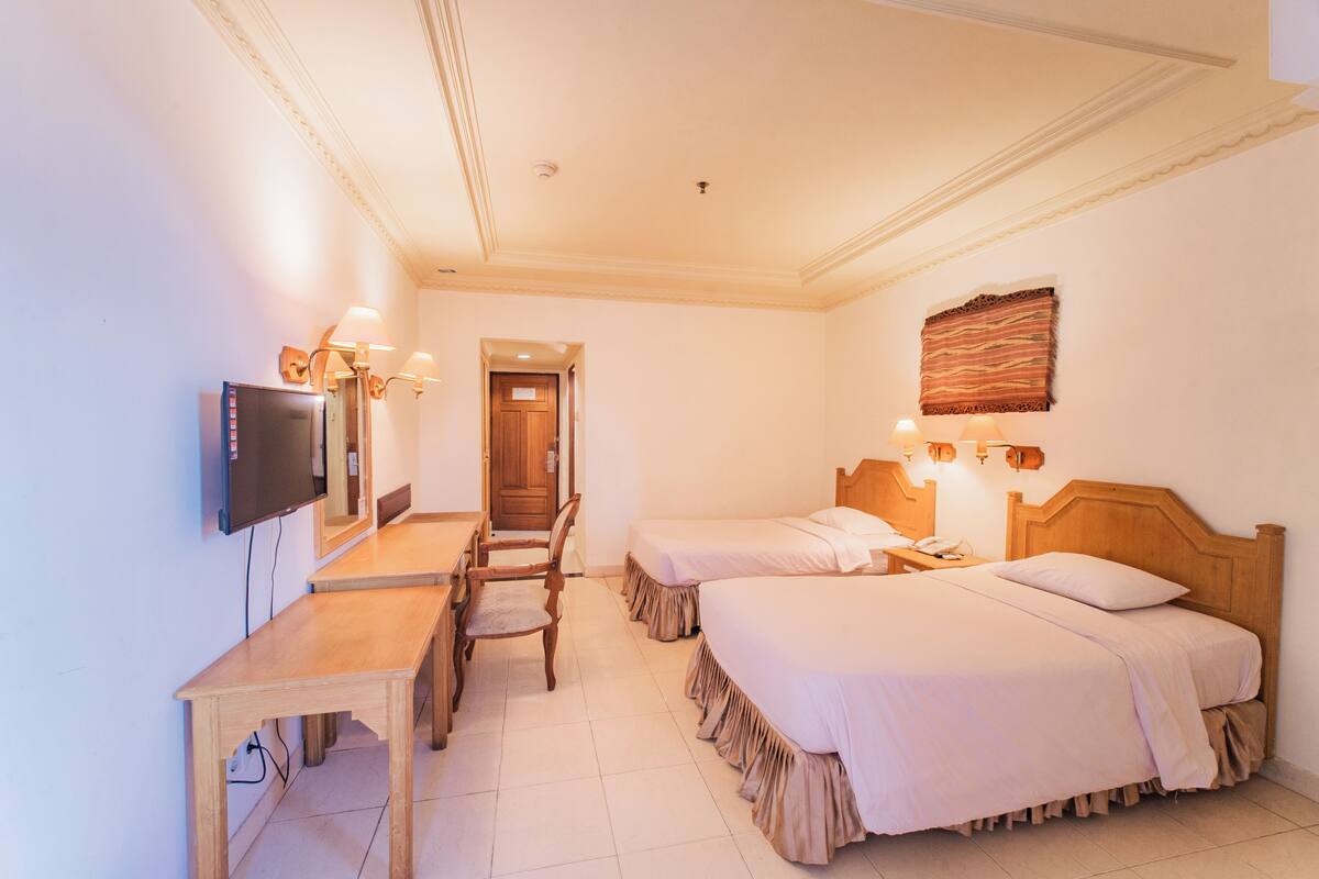 Quarto de hotel com duas camas, uma escrivaninha, uma cadeira, televisão e uma porta que dá para outro quarto.