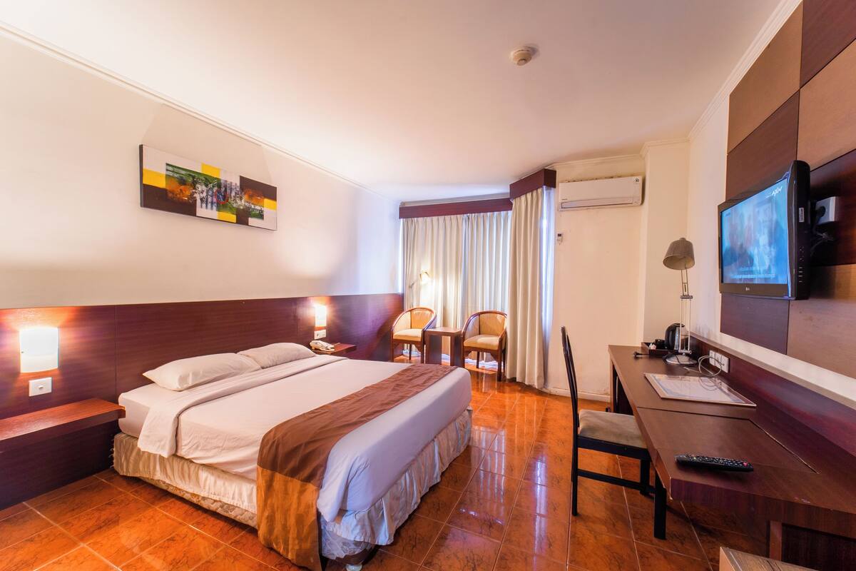 Quarto de hotel com uma cama grande, uma escrivaninha com televisão, uma cadeira e uma mesinha.