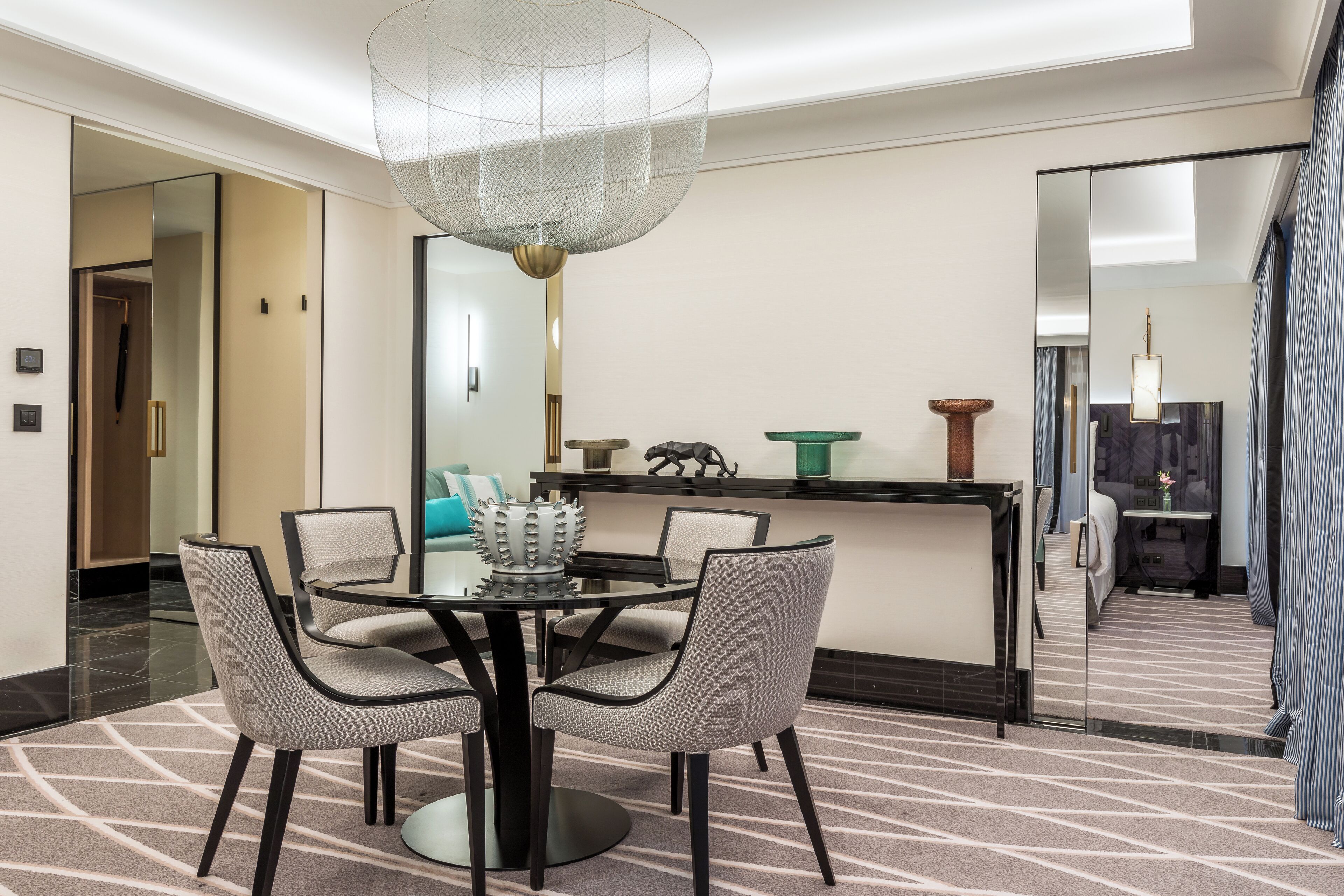 Quarto de hotel moderno com uma mesa de jantar redonda, quatro cadeiras, um candelabro, um espelho grande e vista para o corredor.