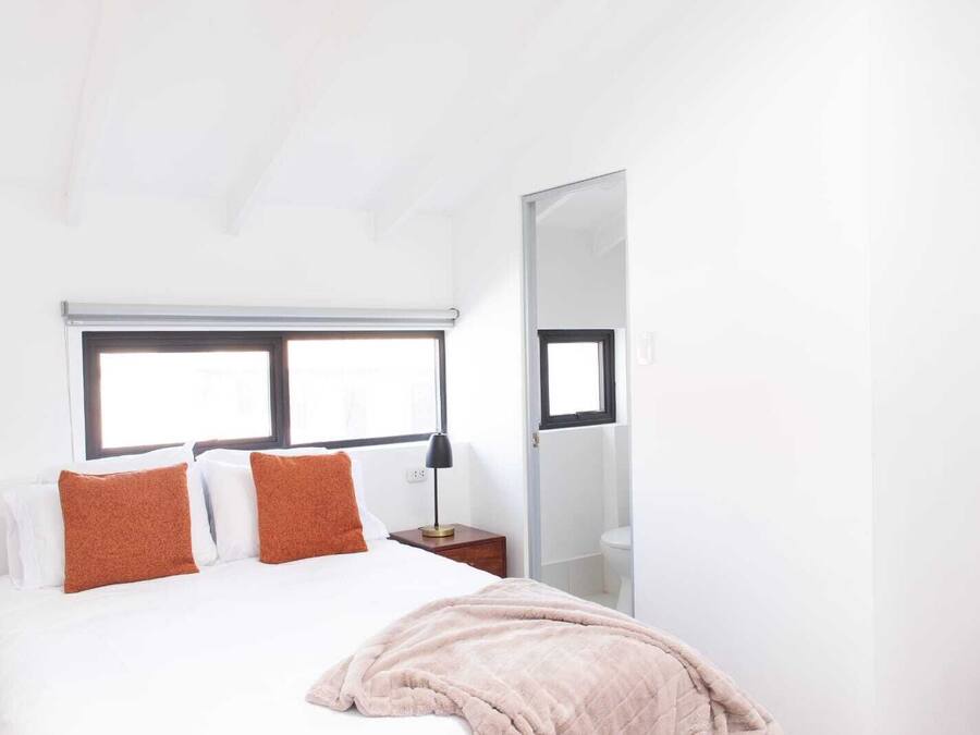 Quarto com uma cama, duas almofadas, uma mesinha de cabeceira com abajur e uma janela.