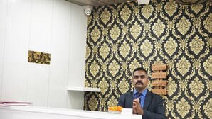 Um homem está atrás de um balcão de recepção em um quarto com papel de parede estampado e um ventilador de teto.