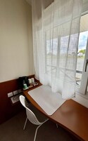 Quarto de hotel pequeno com escrivaninha de madeira, cadeira branca, cafeteira e janela com cortinas finas.