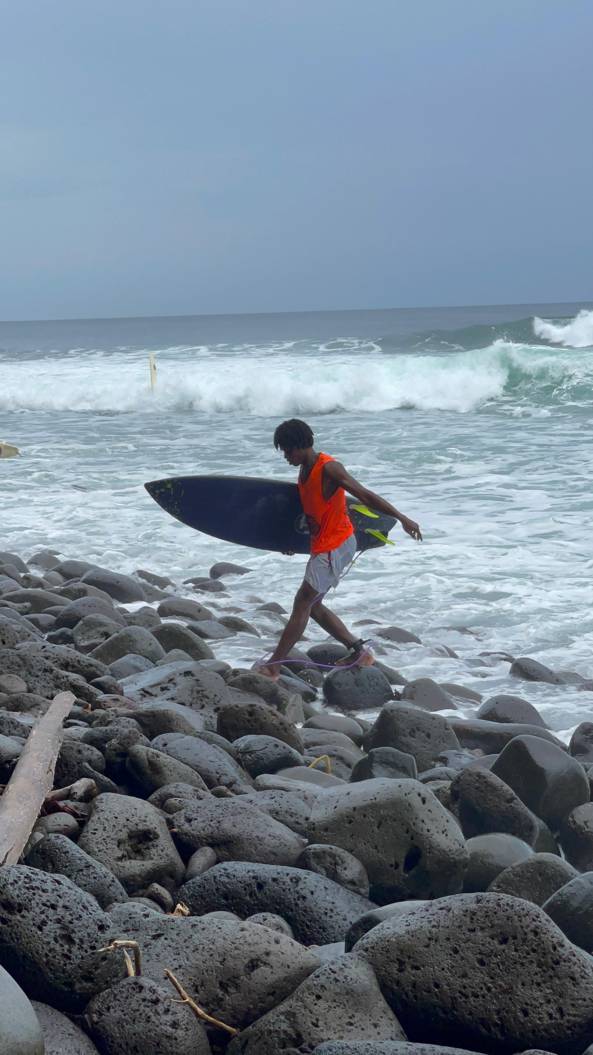 Uma pessoa caminhando numa praia rochosa com uma prancha de surf.