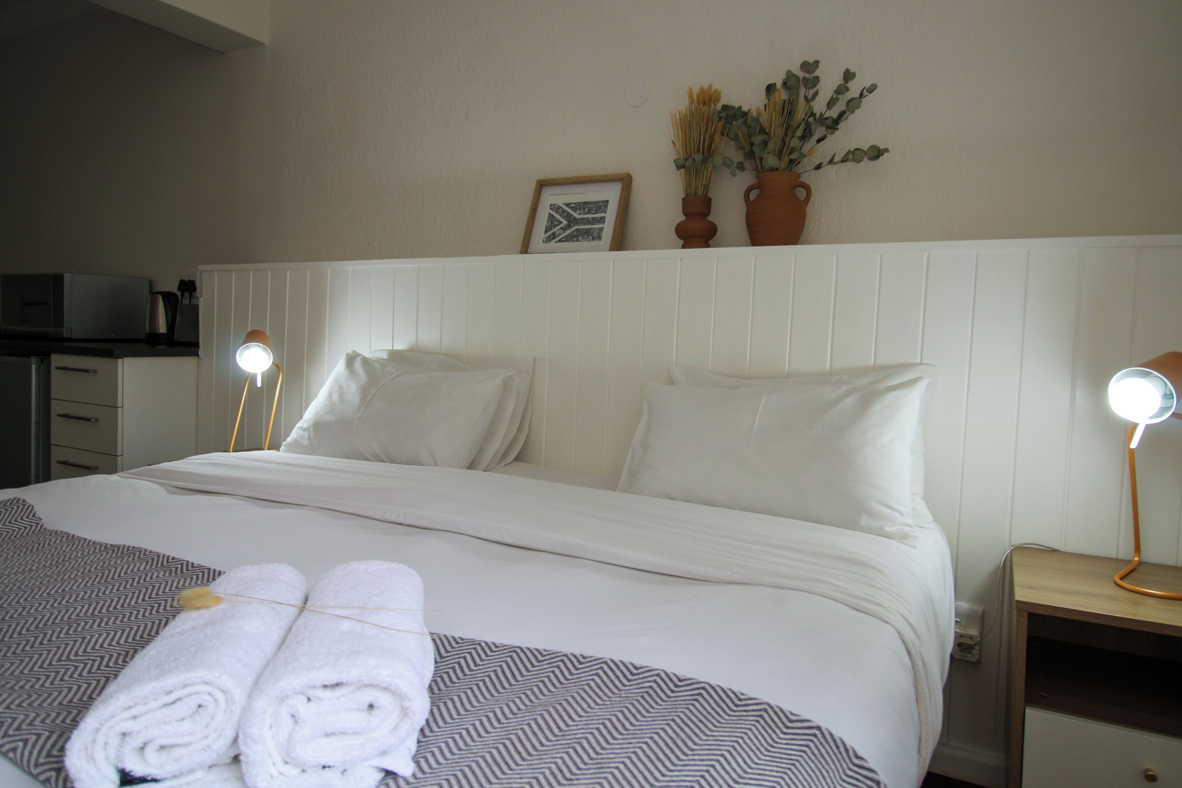 Cama bem arrumada com lençóis brancos, duas luminárias de cabeceira, um quadro emoldurado e um vaso com plantas secas na cabeceira.