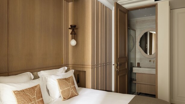 Quarto de hotel com uma cama grande, cabeceira de madeira, luminária fixada na parede e banheiro visível através de uma porta aberta.
