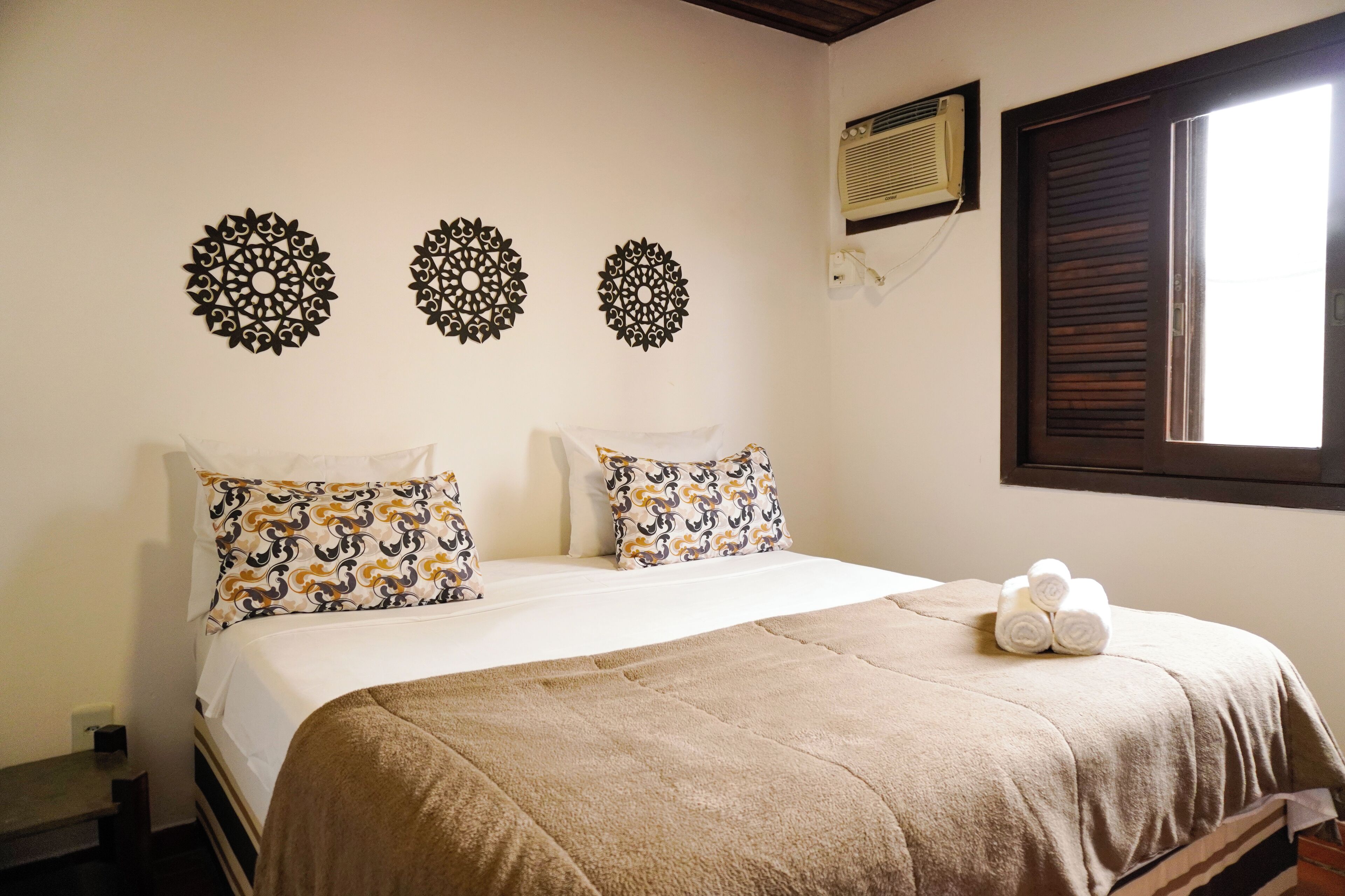 Quarto de hotel com uma cama, duas peças decorativas na parede, um aparelho de ar condicionado e uma janela com persianas.
