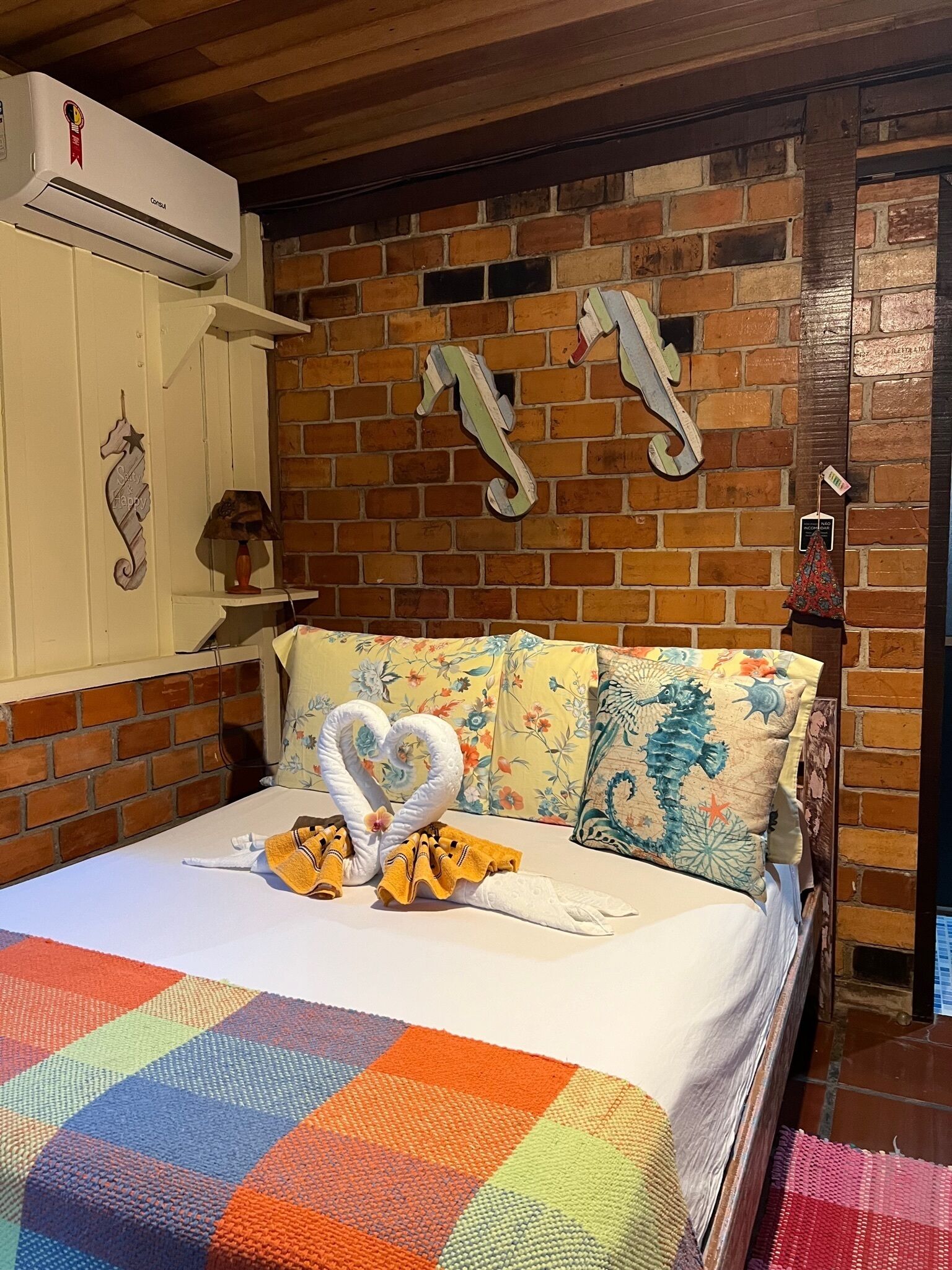 Quarto de hotel com parede de tijolos, cama com almofadas florais, arranjo de toalhas em formato de coração e colcha colorida.